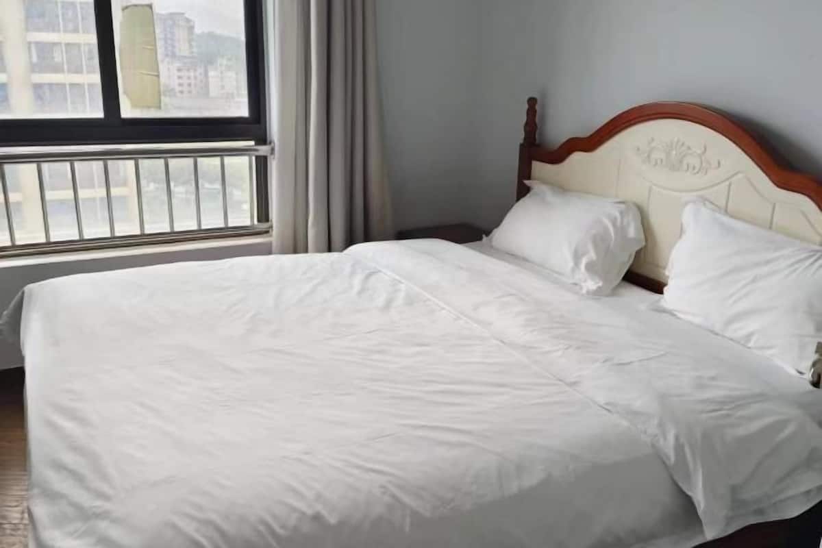 Uma cama com roupa de cama branca, cabeceira de madeira e janela com vista para edifícios.