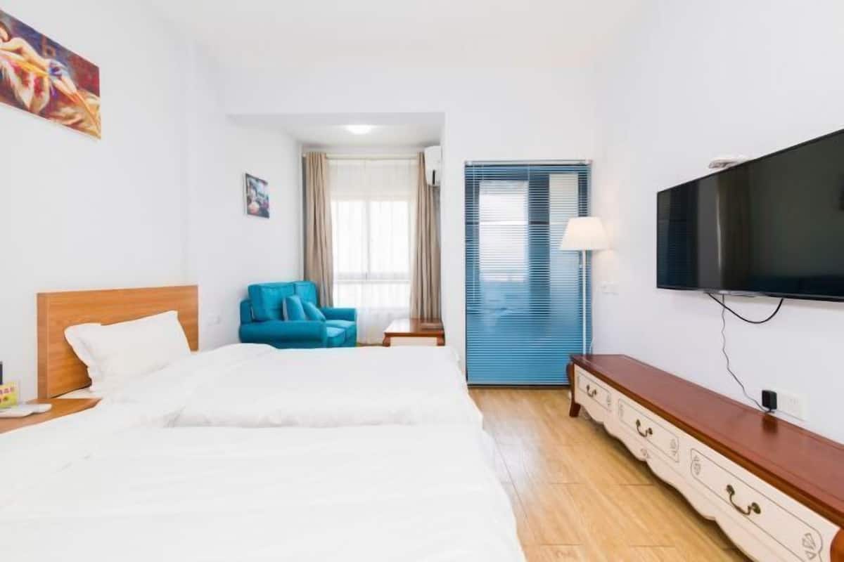 Quarto de hotel com cama, televisão, cadeira azul, cômoda de madeira e porta com persianas.
