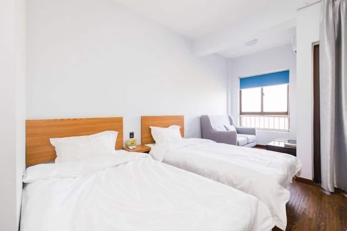 Quarto de hotel com duas camas, uma cabeceira de madeira, uma poltrona cinza e uma janela com vista.