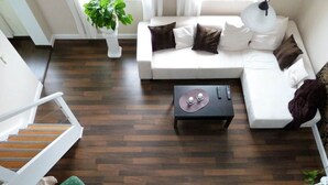 Uma sala de estar moderna com um sofá branco, piso de madeira escura, uma pequena mesa de centro e uma escada com corrimão branco.