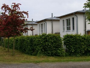 Uma fileira de casas brancas com telhados cinzentos, rodeadas por sebes verdes e uma árvore com folhas vermelhas.