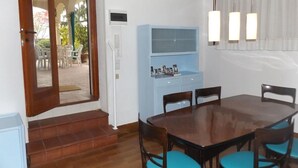 Uma área de refeições com uma mesa e cadeiras de madeira, um armário azul e vista para um pátio externo com cadeiras e plantas.