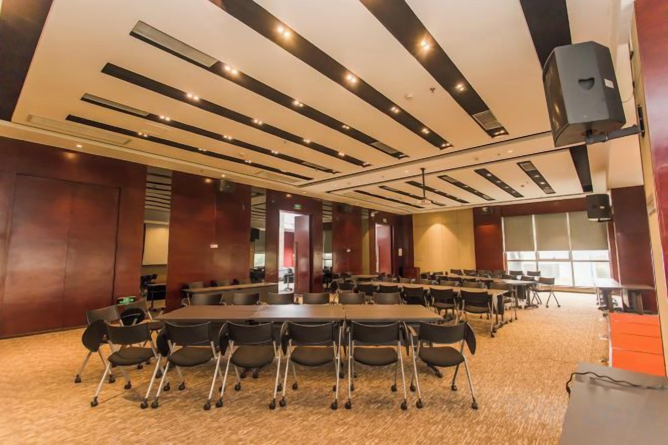 Sala de conferências com várias mesas e cadeiras, teto alto com luminárias e uma grande janela que proporciona iluminação natural.
