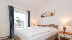 Quarto de hotel com uma cama grande, duas mesas de cabeceira com abajures, uma janela com cortinas e um quadro na parede.