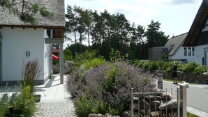 Um edifício com fachada branca e telhado de palha, rodeado por um jardim com flores roxas e um portão de metal.
