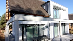 Uma casa moderna com fachada de vidro ampla, um alpendre com mesa e cadeiras, e telhado de palha.
