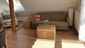 Um espaço de estar acolhedor em estilo loft, com pé-direito alto, vigas de madeira aparentes, um sofá, uma pequena mesa de centro e uma mesinha de vime.