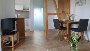 Uma sala de estar moderna com uma mesa de jantar de madeira, duas cadeiras pretas, uma televisão sobre um suporte de madeira, um pequeno armário e um vaso com flores.