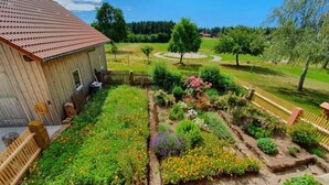 Um jardim com diversas plantas e flores, uma cerca de madeira e um edifício com telhado vermelho.