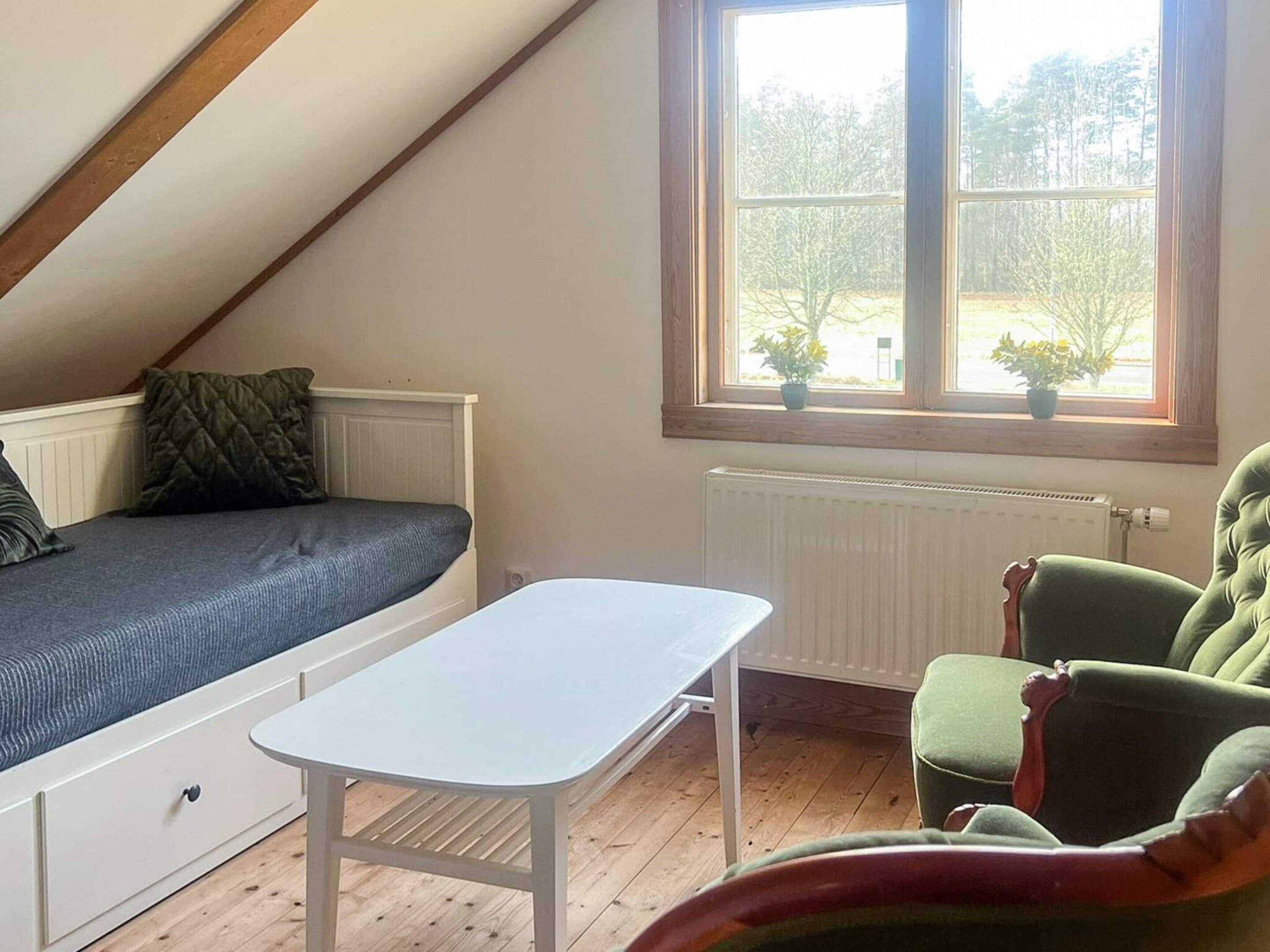 Quarto com teto inclinado, cama com colcha verde, mesa branca, poltrona verde e janela com vista para árvores.