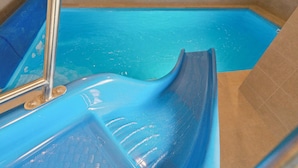 Um escorregador azul em área de piscina, com escada de metal e parede revestida em azulejos.
