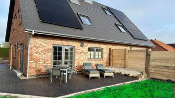 Uma casa com fachada de tijolos, telhado inclinado com painéis solares, garagem e um pátio com móveis de exterior.