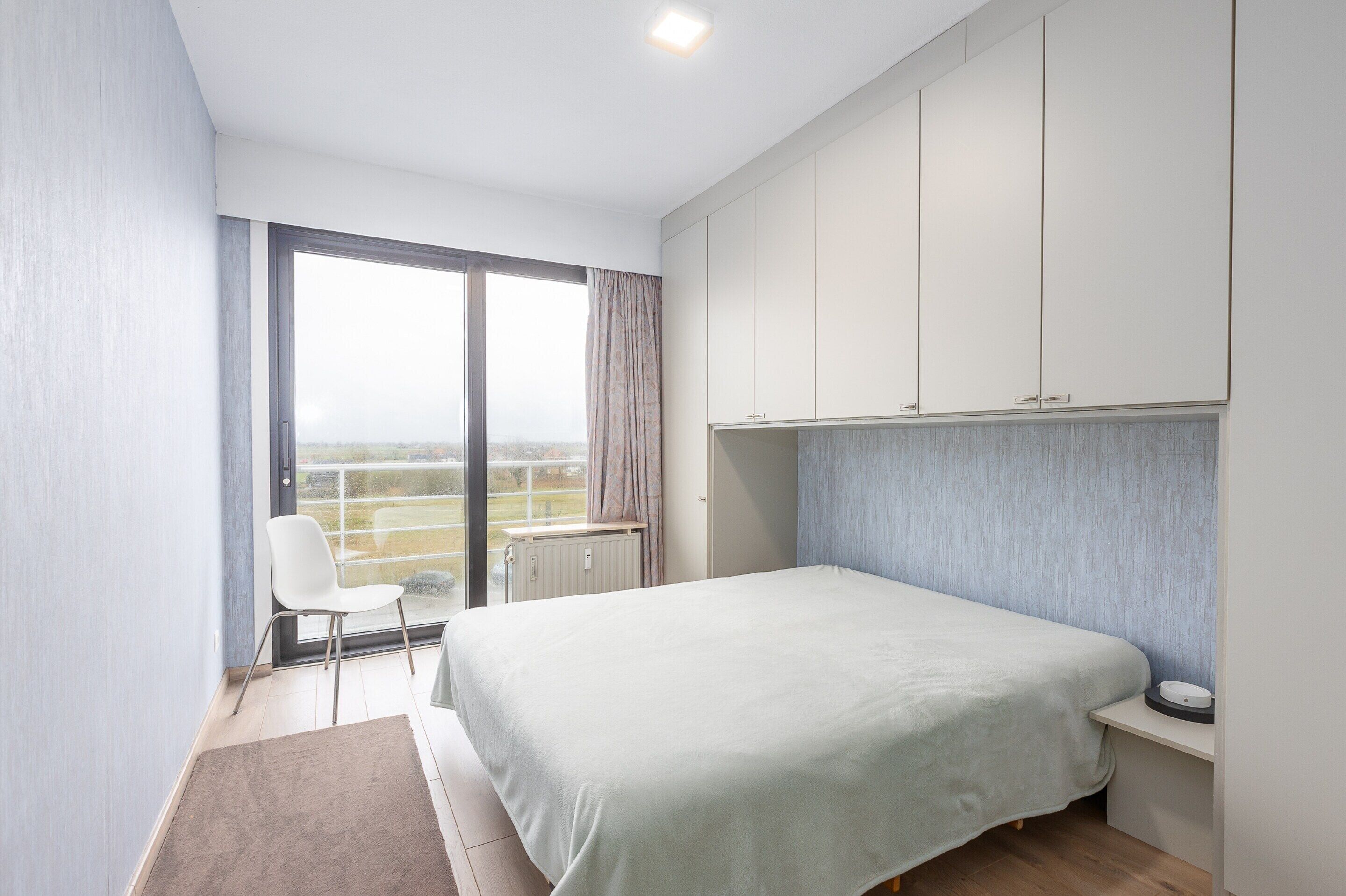 Quarto com cama grande, uma cadeira, janela com cortinas e vista para o exterior.