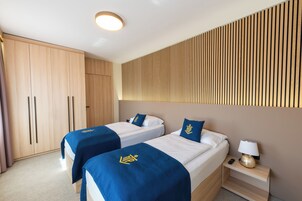 Quarto de hotel com duas camas, um guarda-roupa, uma mesa de cabeceira com abajur e uma parede com linhas verticais.
