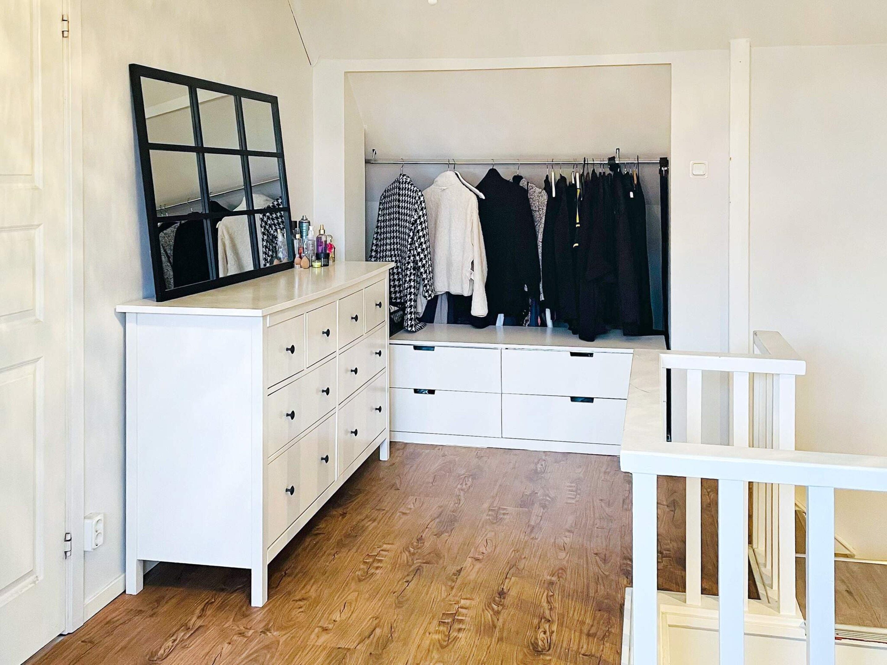 Uma cômoda branca com gavetas, um espelho e um cabideiro com diversas peças de roupa.
