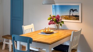 Uma área de jantar com uma mesa de madeira, cadeiras brancas, um vaso com flores, uma luminária pendente e um quadro com uma paisagem na parede.