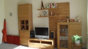 Uma sala de estar com televisão, armários de madeira, uma estante e uma pequena mesa com uma planta.