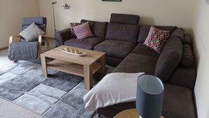Uma sala de estar com um sofá marrom, uma mesa de centro de madeira, uma poltrona cinza e uma almofada colorida.