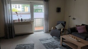 Sala de estar com sofá, mesa de centro e uma poltrona. Há uma janela com cortinas e vista para o exterior.
