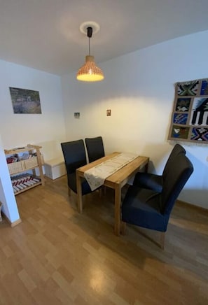 Uma sala de jantar com mesa de madeira e quatro cadeiras, luminária pendente e parede com painel decorativo.