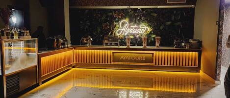Uma área de bar com um letreiro de neon que diz "Ayatoure", piso de mármore polido e balcão de madeira com diversos frascos e copos.