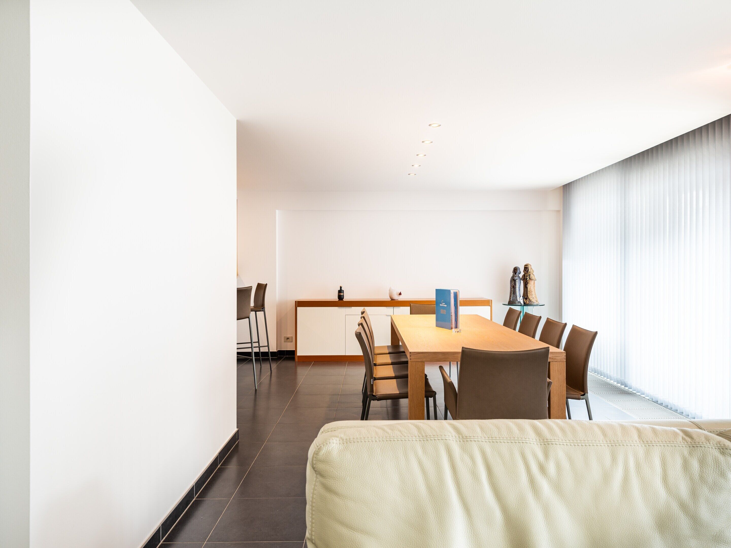 Uma área de refeições moderna com uma mesa e cadeiras de madeira, um pequeno bar com um cardápio azul e uma janela grande com persianas.