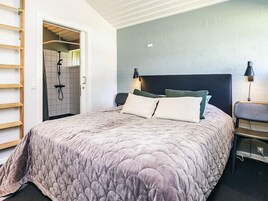 Quarto com uma cama grande, cabeceira, duas luminárias de cabeceira, mesa de cabeceira e uma porta para um banheiro com chuveiro.