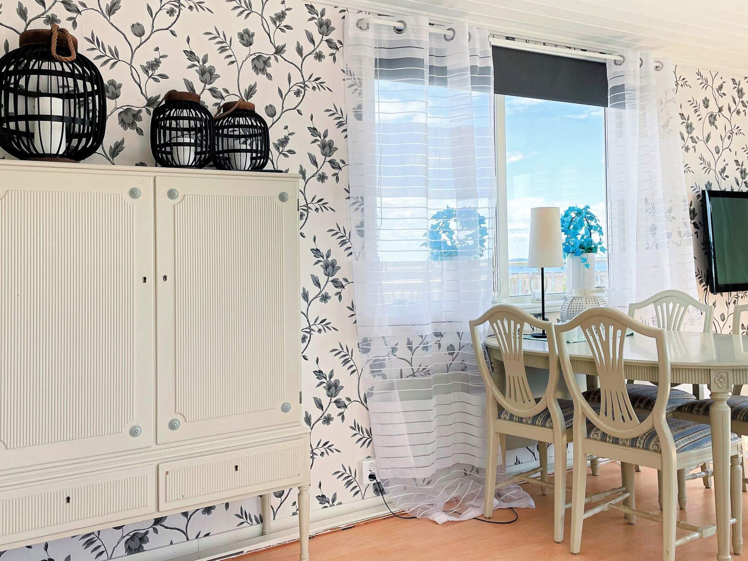 Uma área de refeições com armário branco, papel de parede floral, janela com cortinas leves e uma mesa com cadeiras.