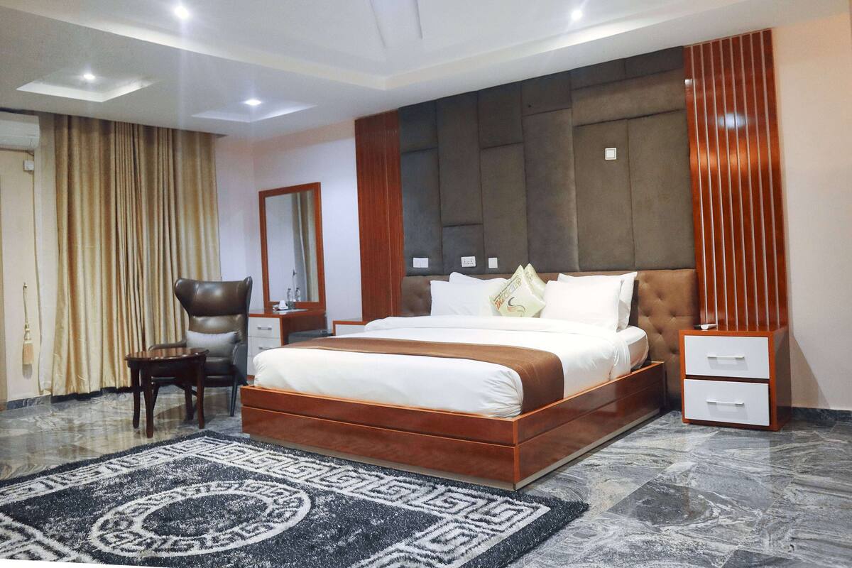 Quarto de hotel moderno com uma cama grande, mesa de cabeceira, cadeira, espelho e um painel decorativo na parede.