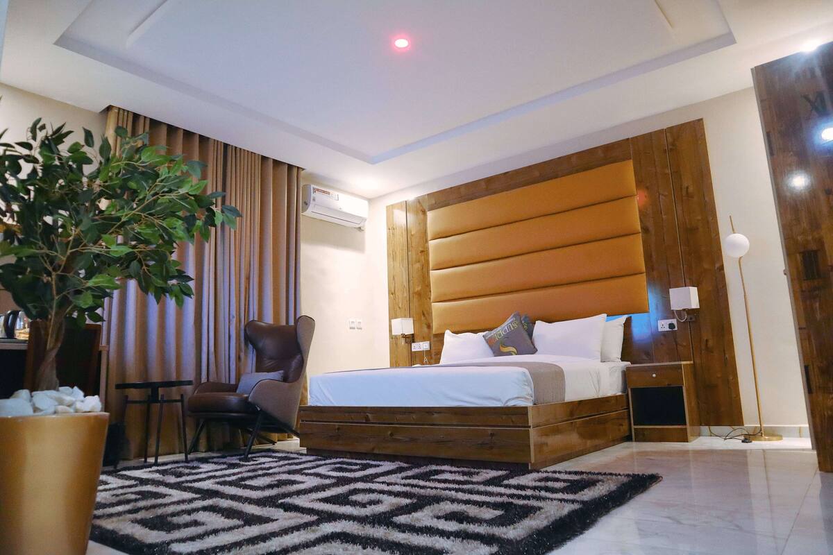 Quarto moderno com cabeceira de madeira, uma cama grande, planta em vaso, uma cadeira e uma mesinha de cabeceira.