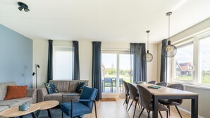 Uma sala de estar moderna com um sofá, uma poltrona azul, uma mesa de centro de madeira e uma mesa de jantar com cadeiras. Há uma janela grande com cortinas, um ventilador de teto e um lustre.