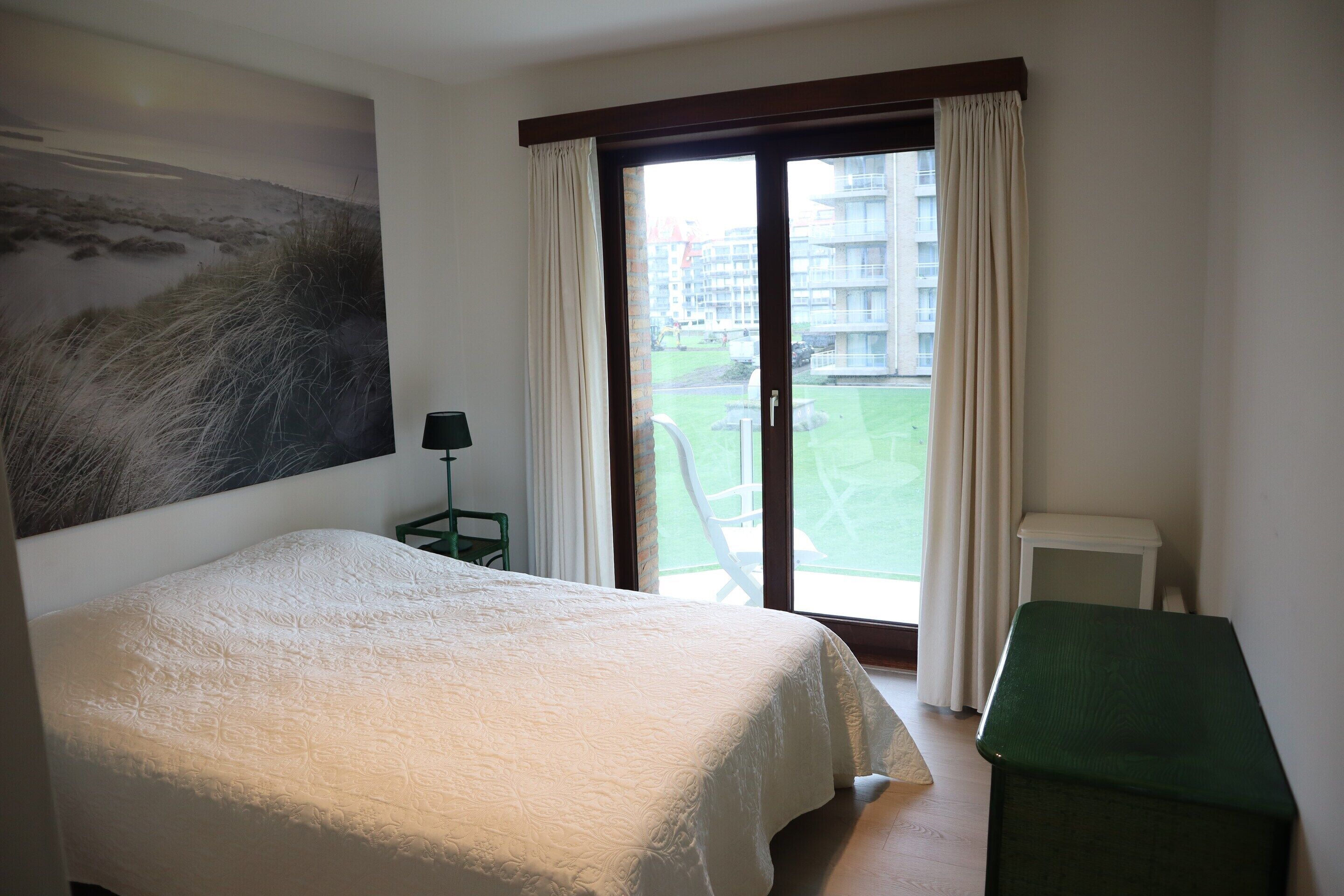 Quarto de hotel com uma cama grande, um quadro na parede, vista para a varanda externa e uma cadeira verde pequena.