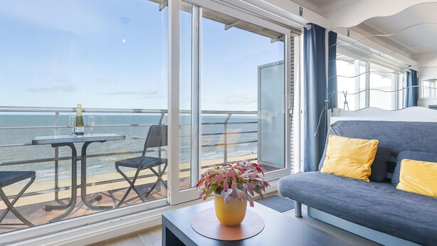 Quarto de hotel moderno com varanda, sofá, mesa com planta e vista para o mar.