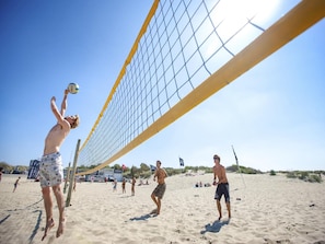 Jogo de voleibol de praia com jogadores de shorts e sem camisa, rede de voleibol e uma praia arenosa com bandeiras e um edifício ao fundo.