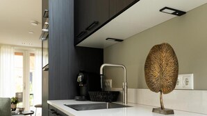Cozinha moderna com bancada branca, armários escuros, cafeteira e uma escultura decorativa em formato de folha.