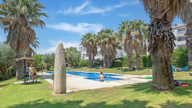 Uma área de piscina com palmeiras, um pilar de pedra e um edifício ao fundo.