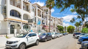 Rua com carros estacionados e edifícios altos com varandas e placas de lojas.