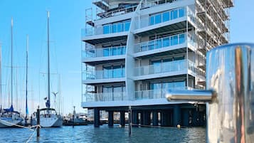 Um edifício com vários andares e varandas, uma marina com barcos, e um céu limpo.