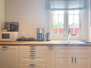 Cozinha com micro-ondas, cafeteira e chaleira no balcão, janela com vista para um edifício vermelho e pia com torneira.
