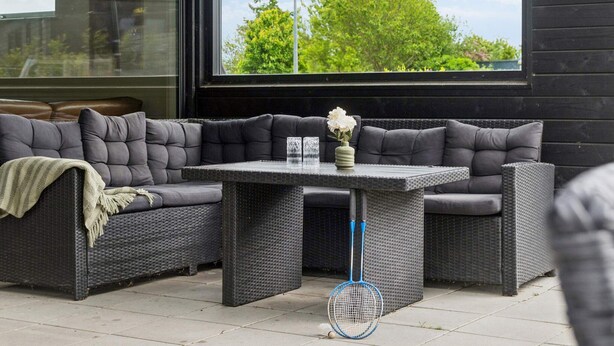 Uma área de estar externa com um sofá escuro e uma mesa, uma raquete de badminton e um vaso com flores.