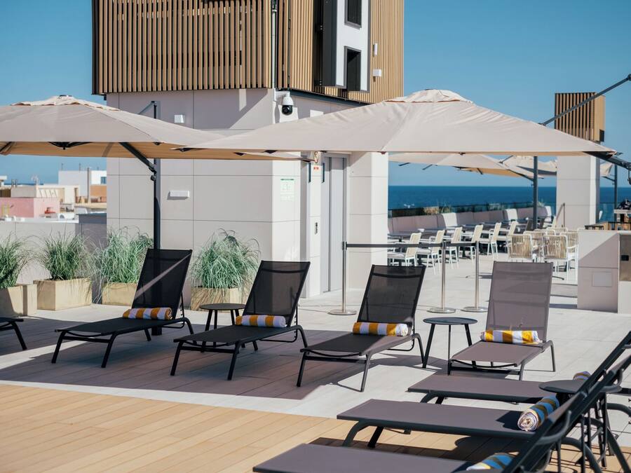 Terraço no telhado com espreguiçadeiras e guarda-sóis, com vista para a paisagem urbana e o mar.