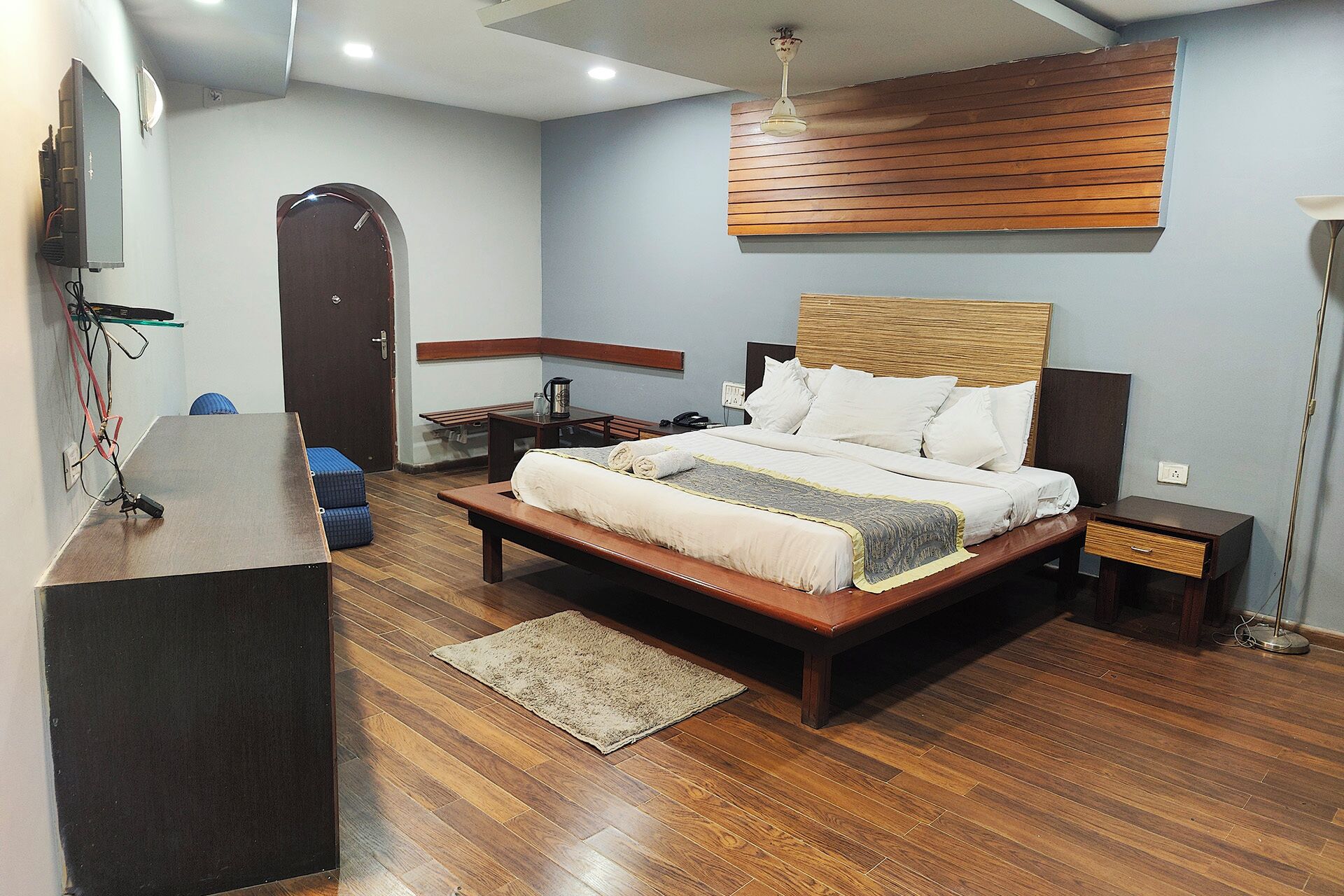 Um quarto com cama de madeira, uma escrivaninha, uma cadeira, um banquinho, uma pequena mesa com chaleira e uma mesinha de cabeceira com abajur.