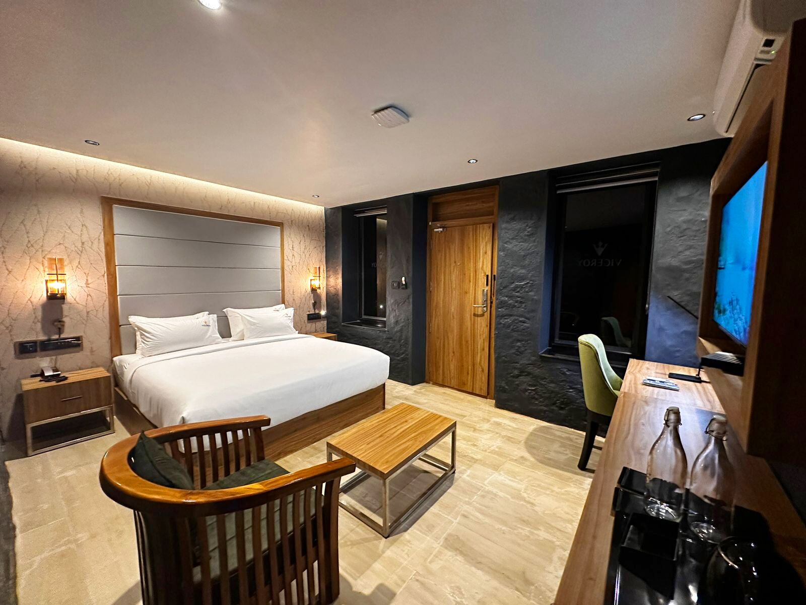 Quarto de hotel moderno com uma cama grande, uma cadeira de madeira, uma escrivaninha com computador e televisão.