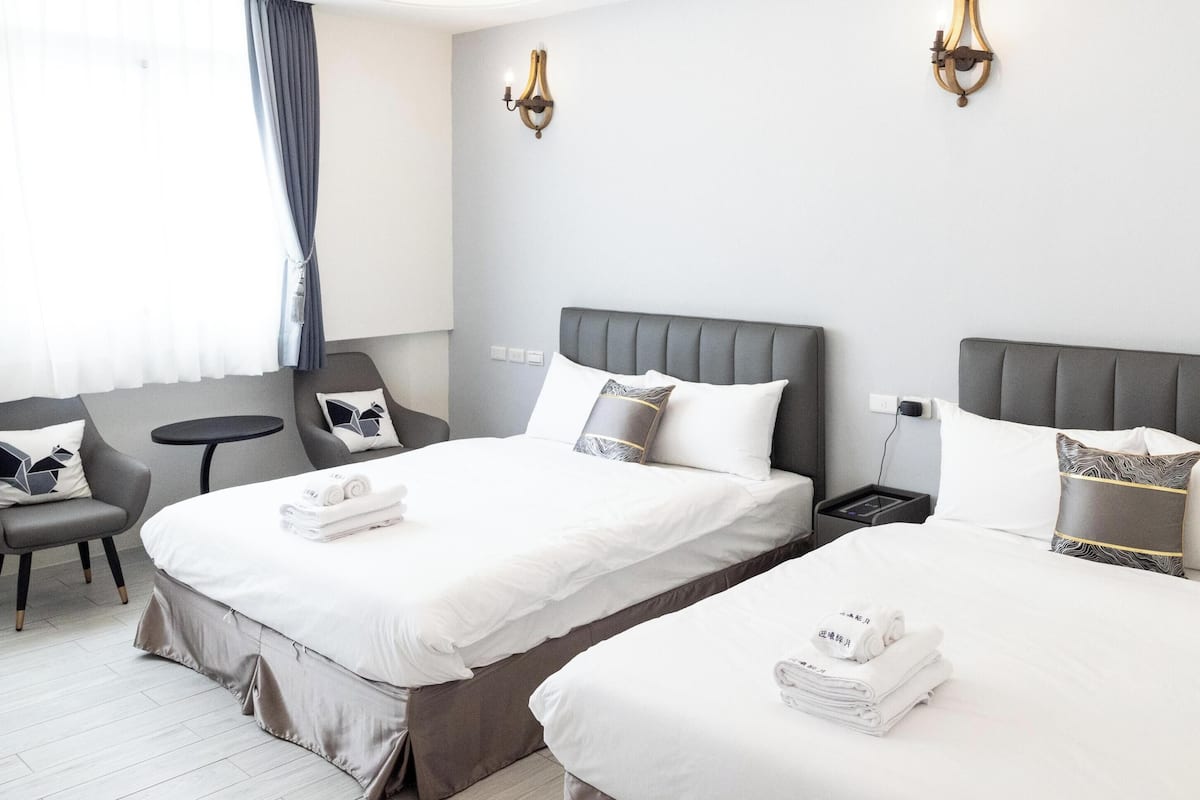 Quarto de hotel com duas camas, uma cadeira, uma mesinha e uma mesa de cabeceira.
