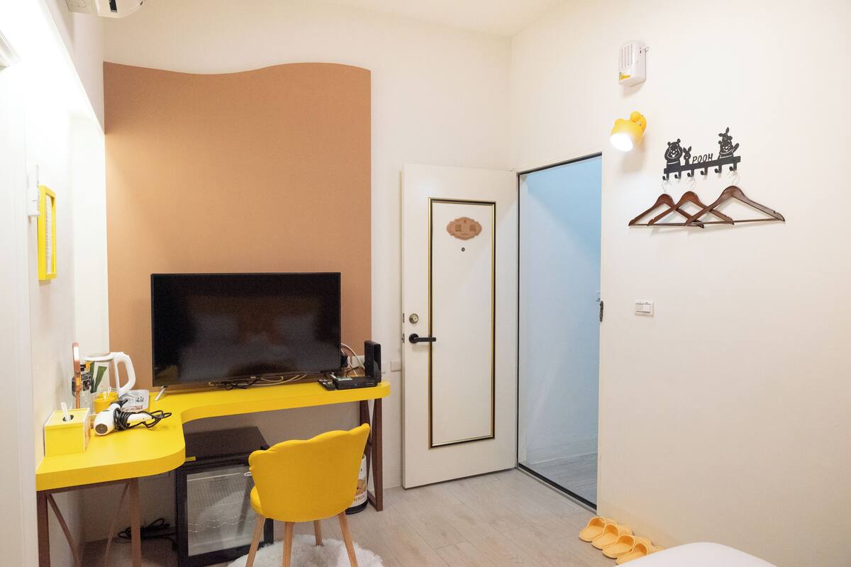 Quarto compacto com escrivaninha amarela, televisão, mini-geladeira e porta para uma varanda.
