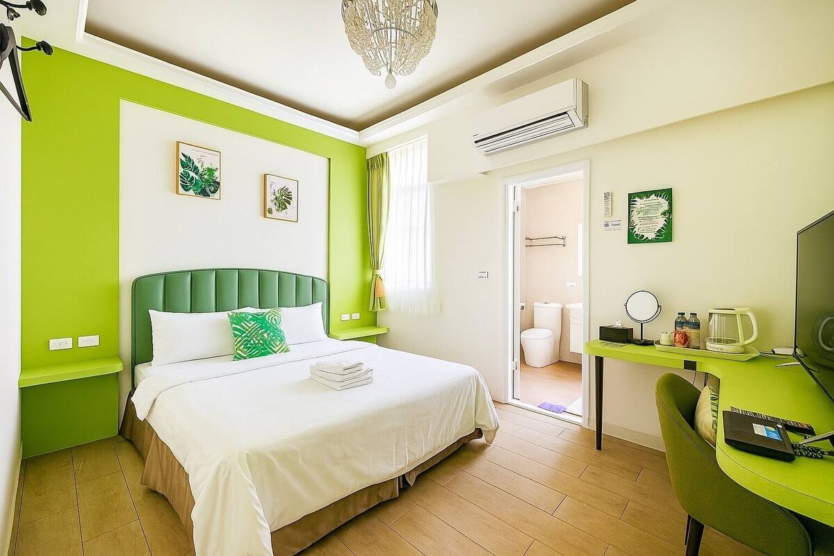 Quarto de hotel com cama, escrivaninha com computador, televisão e banheiro visível através de uma porta aberta.
