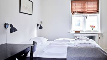 Quarto de hotel com uma cama, duas luminárias de cabeceira, uma escrivaninha, um radiador e uma janela com vista para edifícios.