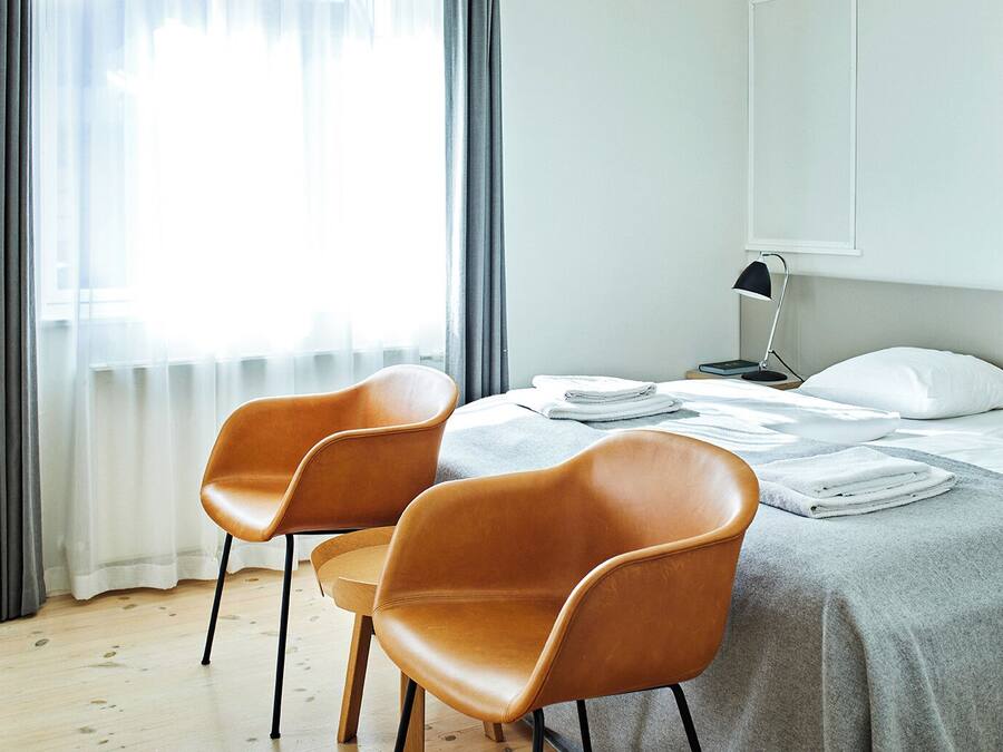 Quarto de hotel com cama, duas cadeiras, mesa de cabeceira com abajur e janela com cortinas.
