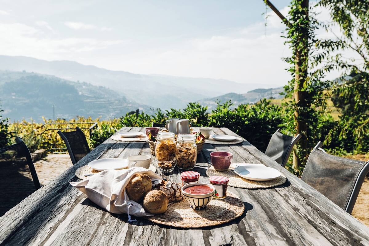 Mesa de madeira posta ao ar livre com diversos alimentos e bebidas, rodeada por cadeiras, com uma vista panorâmica de montanhas ao fundo.