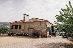Um edifício de pedra com telhado de telhas, uma chaminé e uma pequena escadaria que leva a uma porta. Há vasos de flores com flores cor-de-rosa e uma árvore à direita.